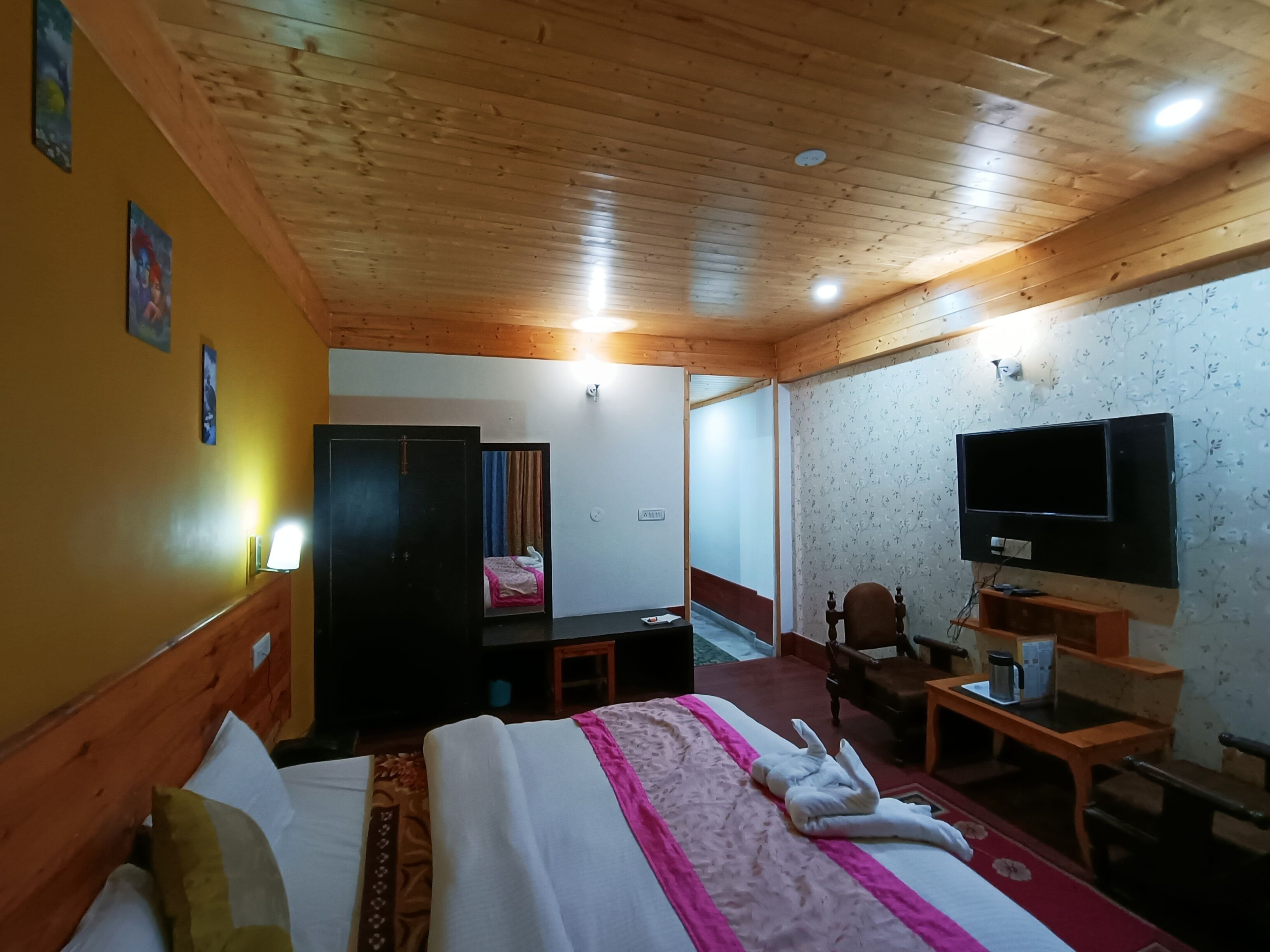 Quarto de hotel com tetos de madeira, uma cama com manta rosa e branca, uma televisão na parede e uma pequena mesa com chaleira.