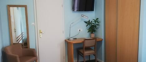 Quarto de hotel com cama, escrivaninha com cadeira, televisão fixada na parede, planta em vaso e guarda-roupa.