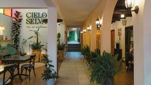 Corredor de hotel com área de refeições, plantas em vasos e escada que leva à entrada.