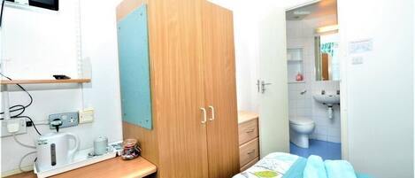 Quarto com cama, guarda-roupa de madeira, escrivaninha com chaleira, televisão e banheiro visível por uma porta aberta.