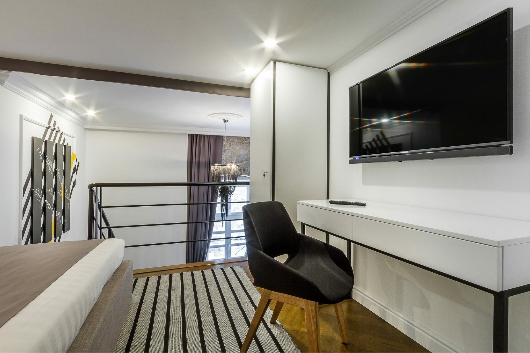 Quarto de hotel moderno com uma televisão de tela plana grande, uma cama com cabeceira de madeira, uma cadeira preta e um tapete listrado.