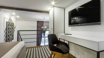 Quarto de hotel moderno com uma televisão de tela plana grande, uma cama com cabeceira de madeira, uma cadeira preta e um tapete listrado.