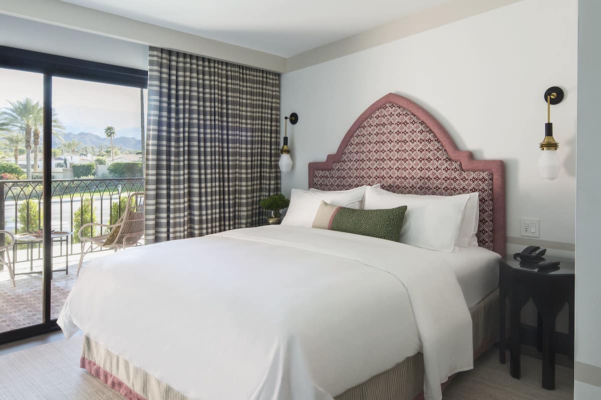 Quarto de hotel com uma cama grande, uma varanda com cortinas, uma mesa pequena e uma mesa de cabeceira com uma luminária.