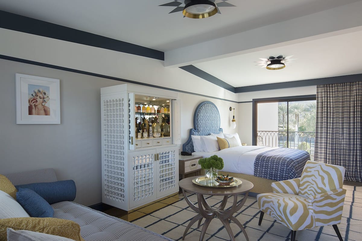 Quarto de hotel com cama, área de estar com sofá e poltronas, minibar e uma janela grande com vista.