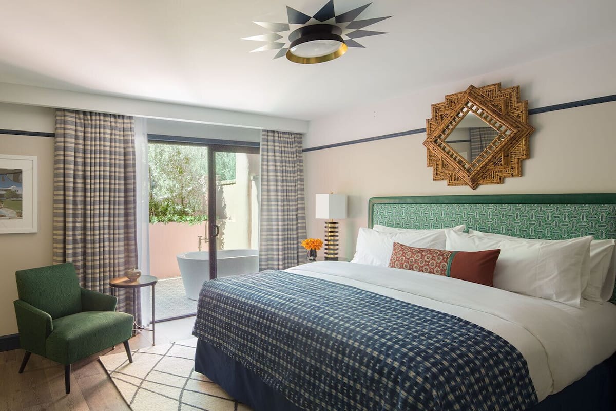 Quarto de hotel com uma cama, uma poltrona verde, uma mesinha de cabeceira com abajur e uma janela grande com cortinas.