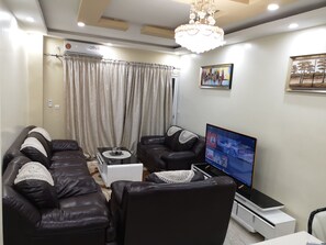Uma sala de estar moderna com um sofá seccional, uma TV de tela plana, uma mesa de centro e um lustre.