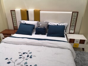 Cama bem arrumada com colcha branca e azul, várias almofadas e mesa de cabeceira de madeira com gaveta.