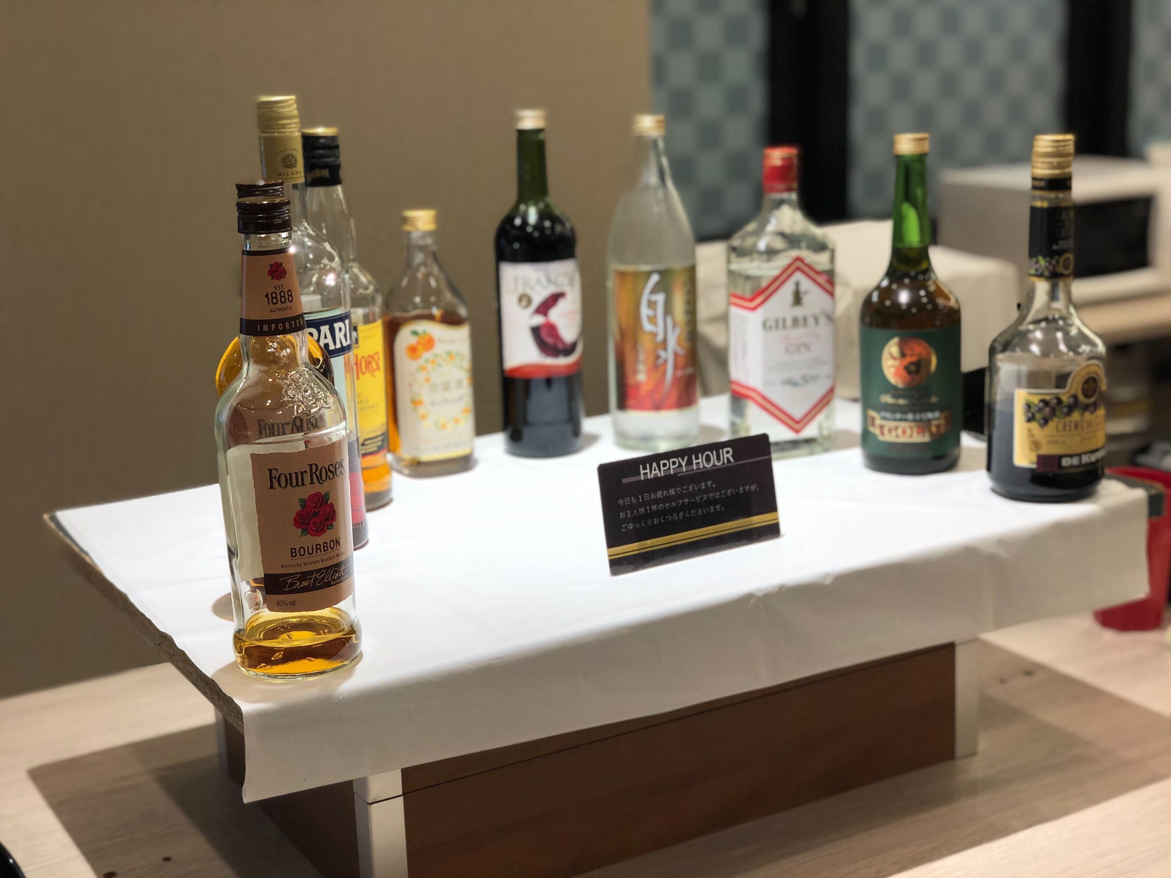Vários garrafas de bebidas alcoólicas sobre uma mesa, com um aviso que diz 'Happy Hour'.