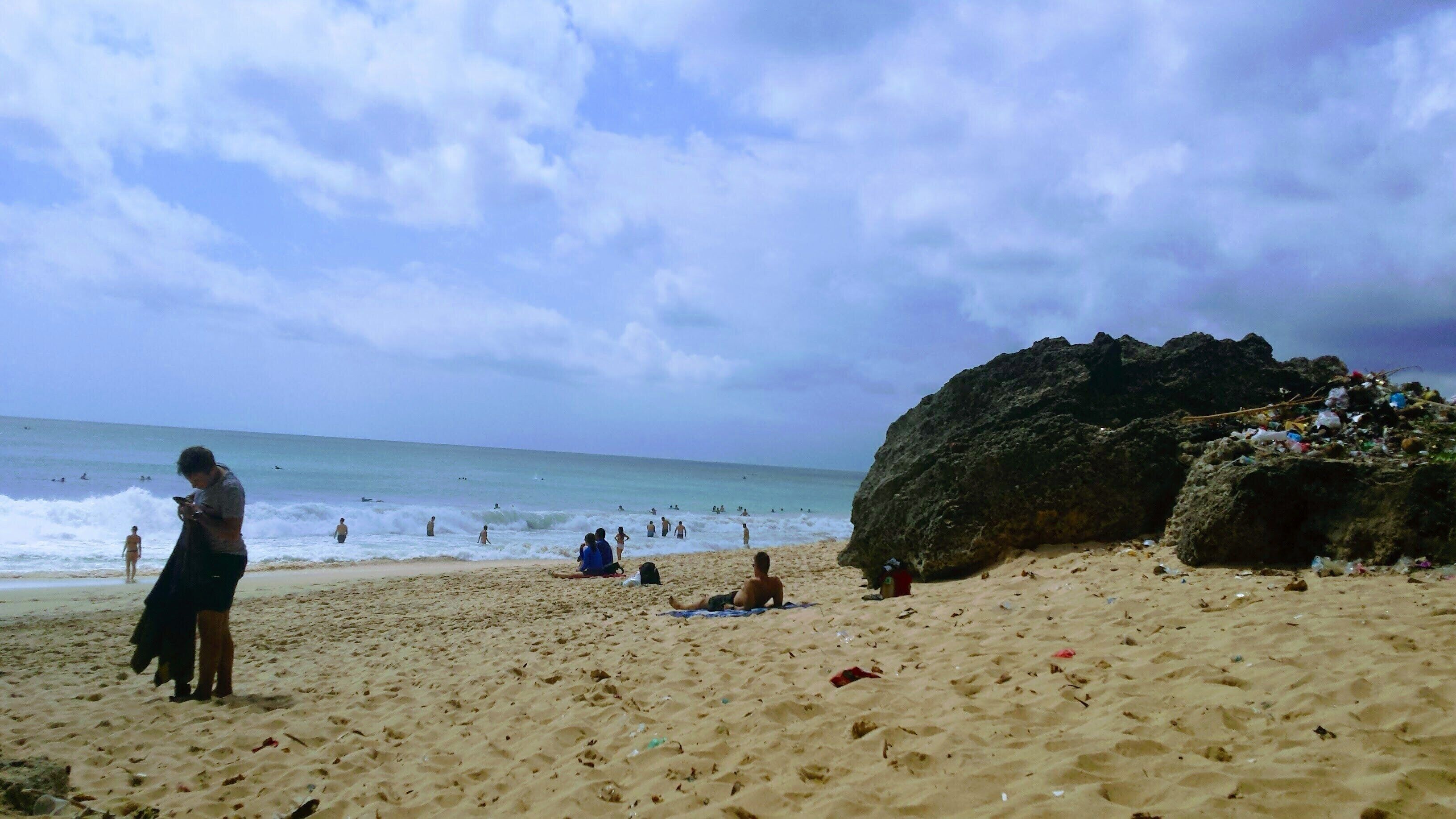 Uma praia arenosa com uma grande formação rochosa, ondas e pessoas na água.