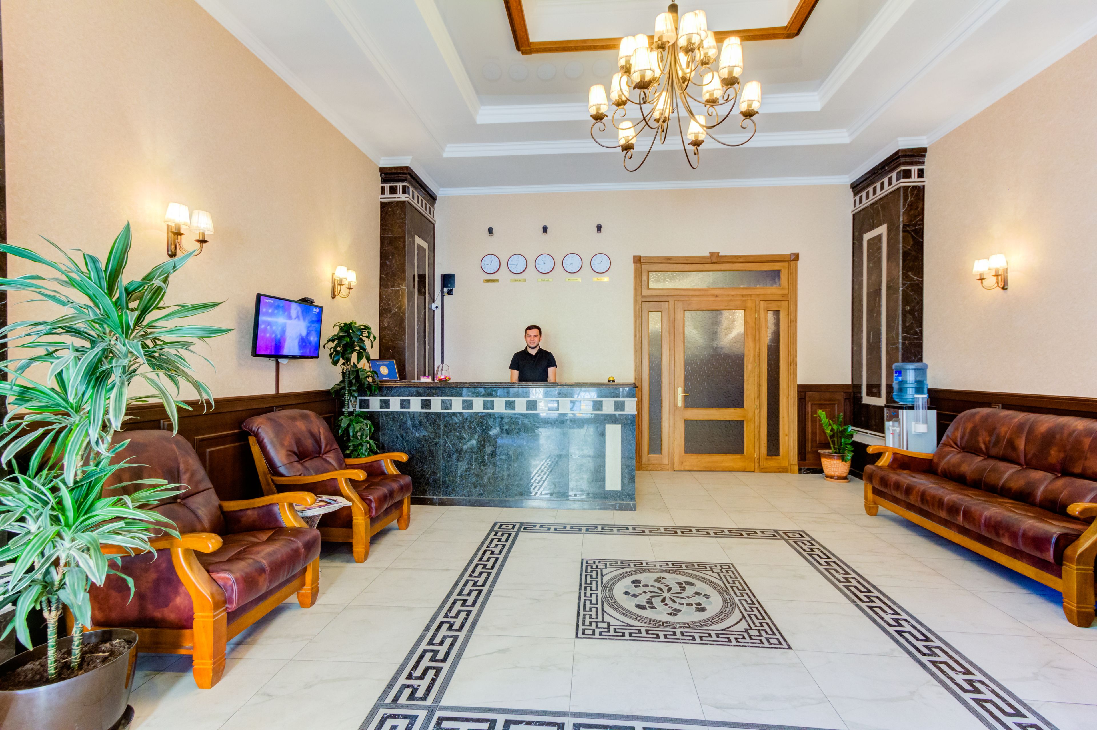 Um saguão de hotel com balcão de recepção, sofás de couro, um lustre, uma televisão e um tapete decorativo.