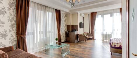 Quarto de hotel espaçoso com piso de madeira, mesa de centro de vidro, sofá, cama, TV e varanda com cortinas.