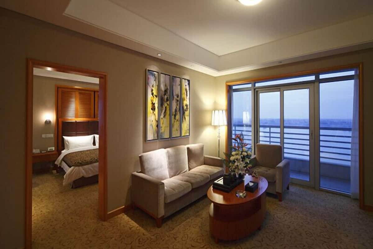 Quarto de hotel com cama, sofá, mesa de centro e uma janela grande com vista para o mar.