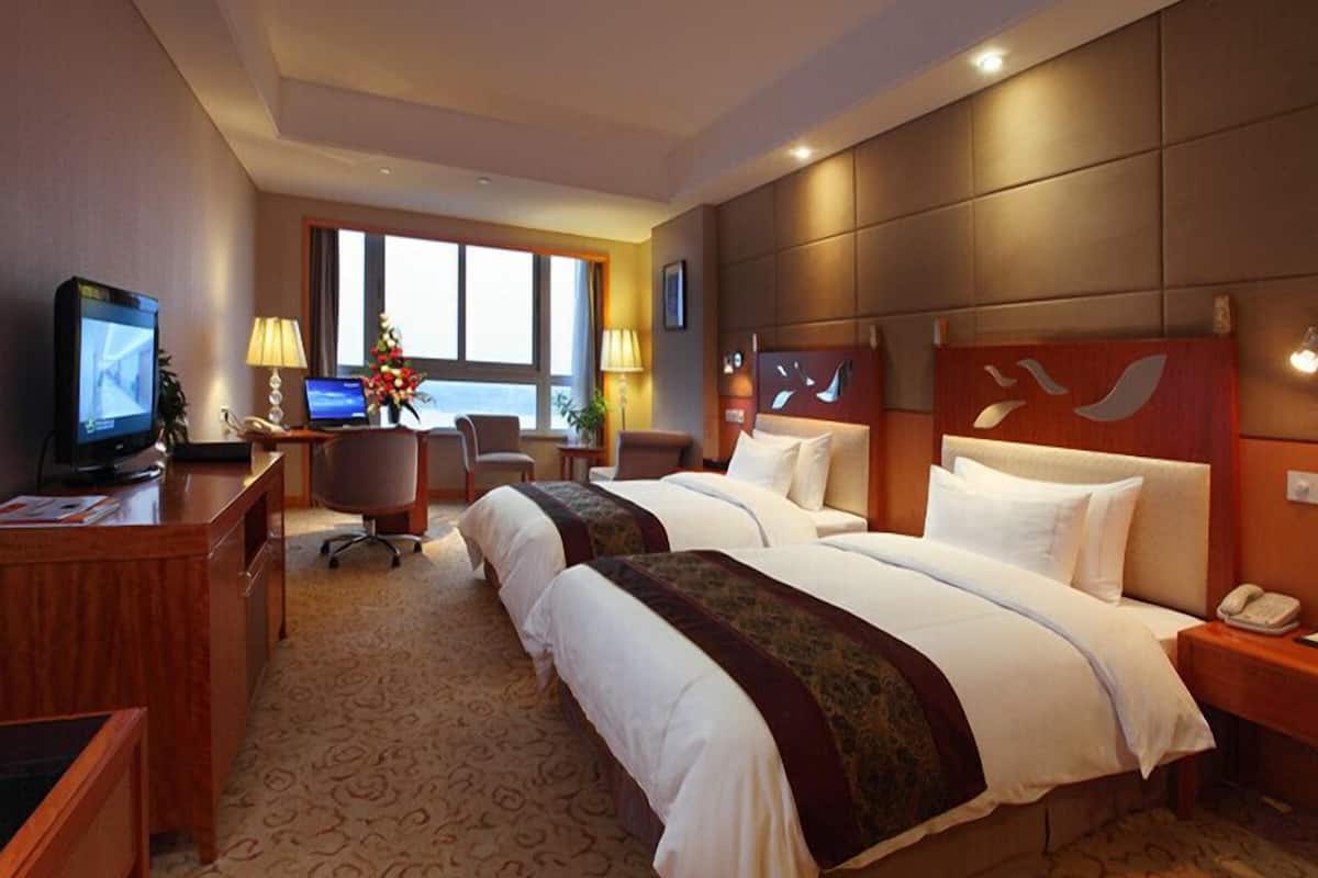 Quarto de hotel com duas camas, uma mesa com computador, televisão e janela com vista.