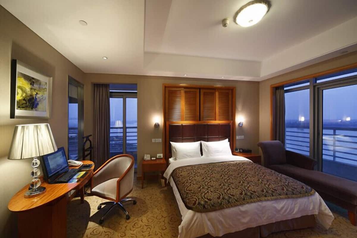 Quarto de hotel com uma cama grande, uma escrivaninha com um laptop, uma cadeira e vista para o mar através de janelas amplas.