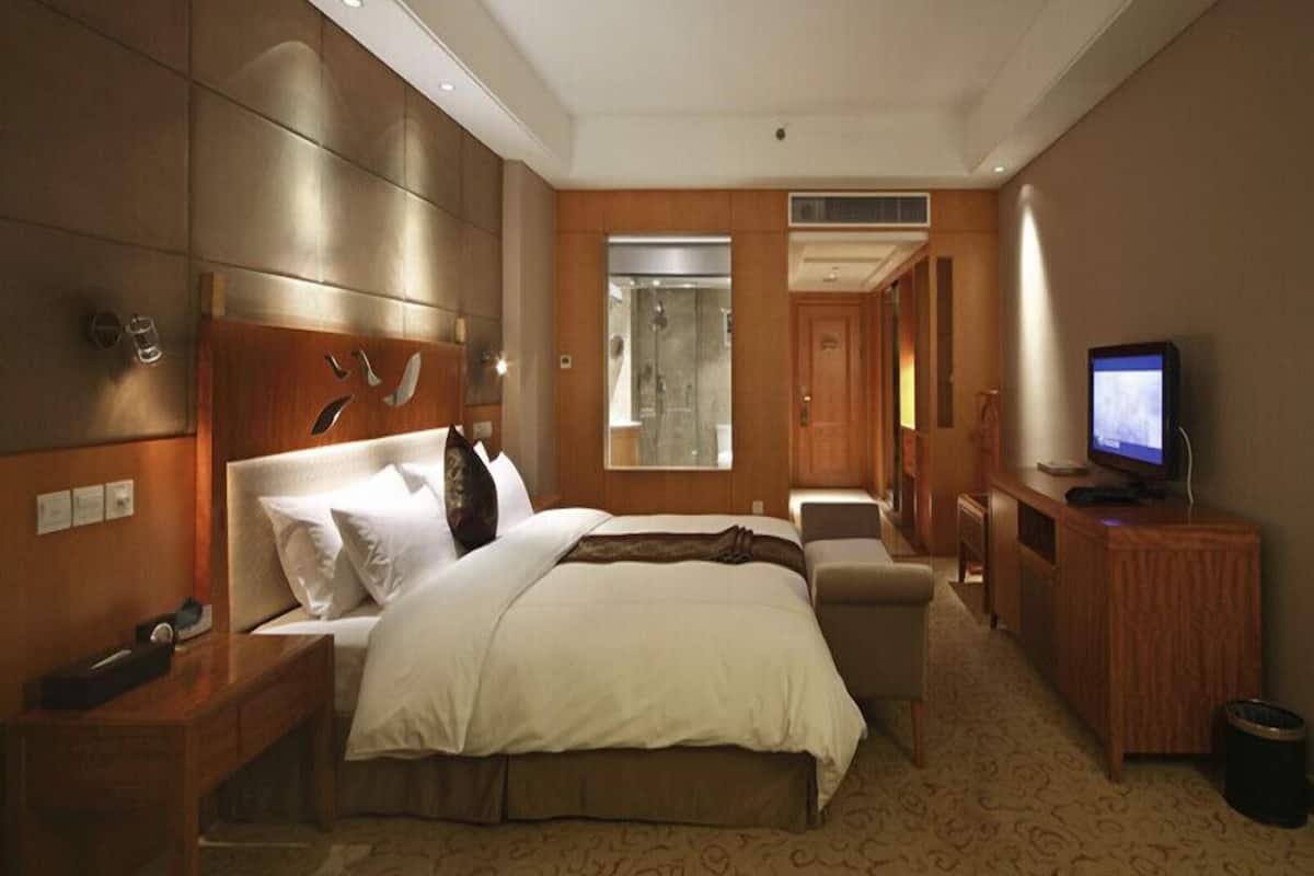Quarto de hotel com uma cama grande, uma mesa com computador, televisão e banheiro visível por uma porta de vidro.