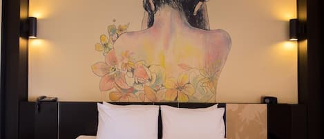 Quarto de hotel com uma cama grande, duas luminárias de cabeceira e um mural na parede retratando uma mulher com flores.