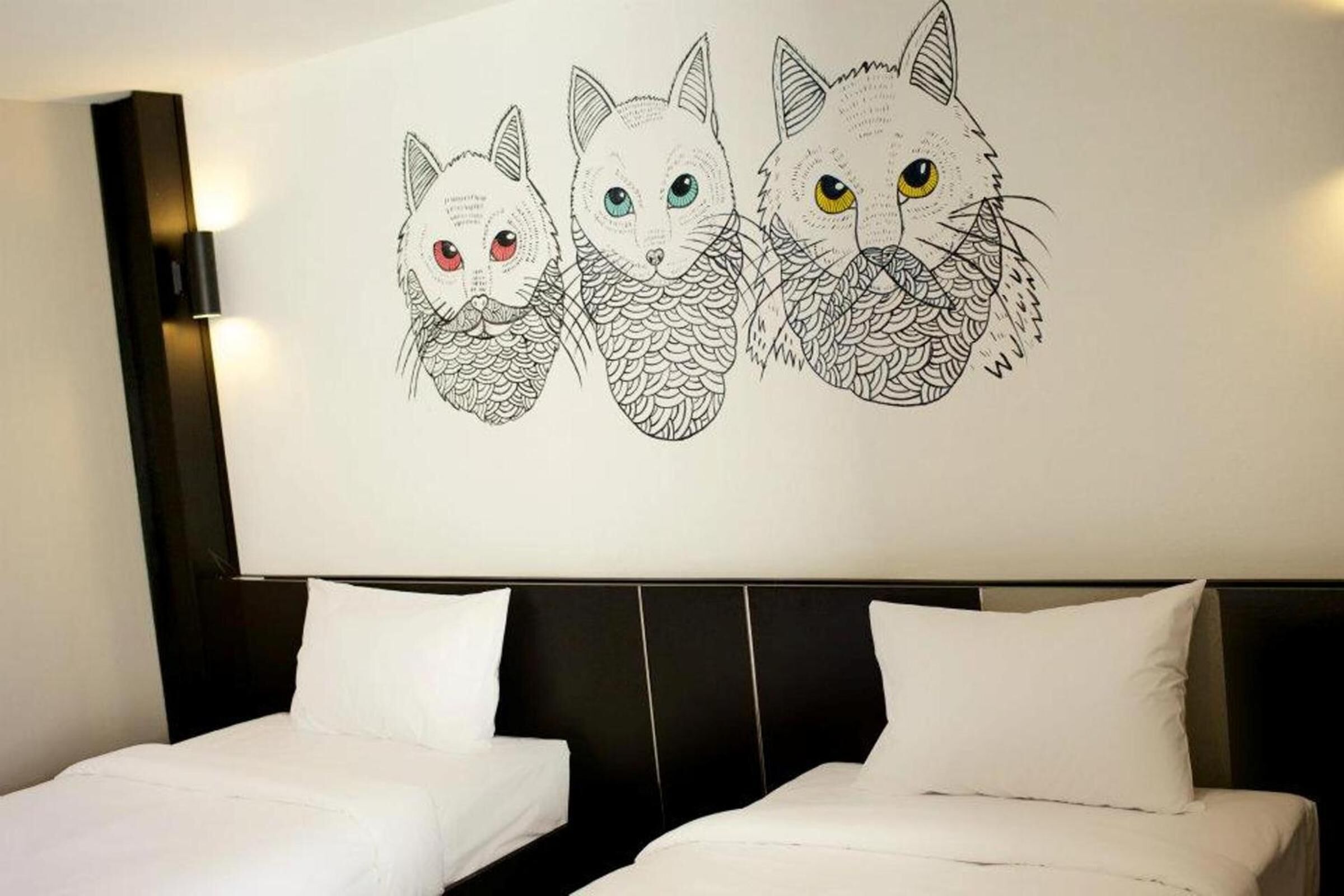 Quarto de hotel com duas camas, uma cabeceira escura e uma parede com um desenho grande e detalhado de três gatos.