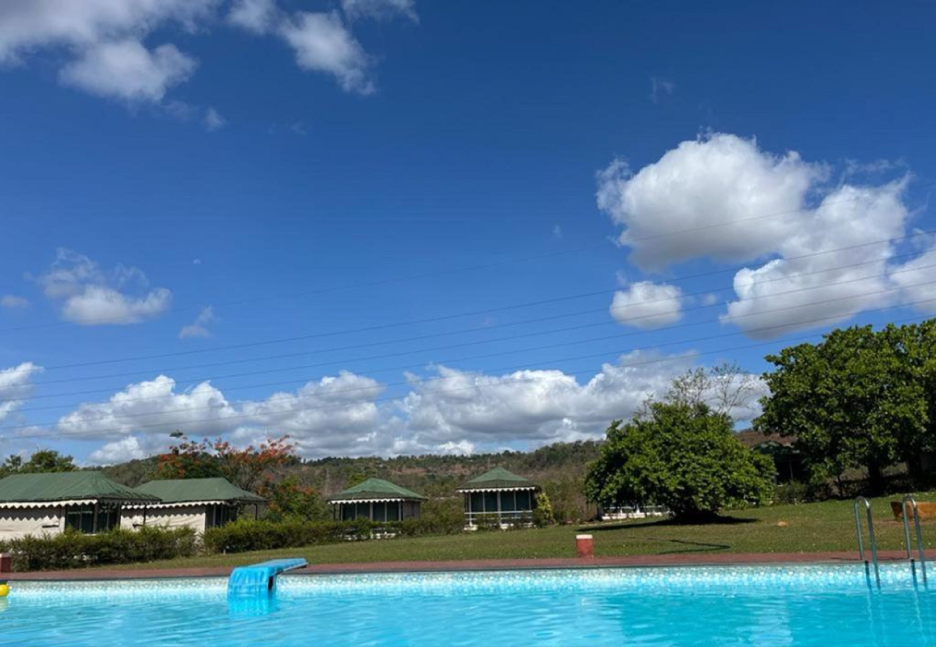 Piscina sob um céu azul límpido com nuvens, rodeada por árvores verdes e um edifício com telhado verde.