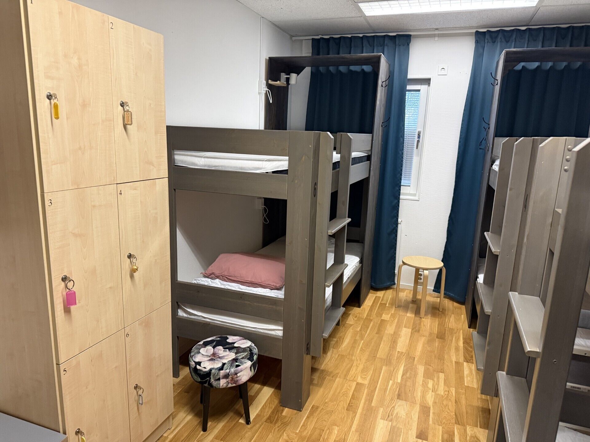 Quarto de dormitório com beliches, guarda-roupa de madeira, um banquinho pequeno e uma janela com cortinas.