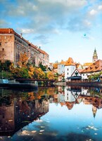 Um edifício histórico com uma vasta superfície aquática refletora à frente, rodeado por árvores de outono e arquitetura tradicional.