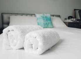 Cama bem arrumada com toalhas brancas dobradas e um travesseiro azul-turquesa com a inscrição 'SEAS THE DAY'.
