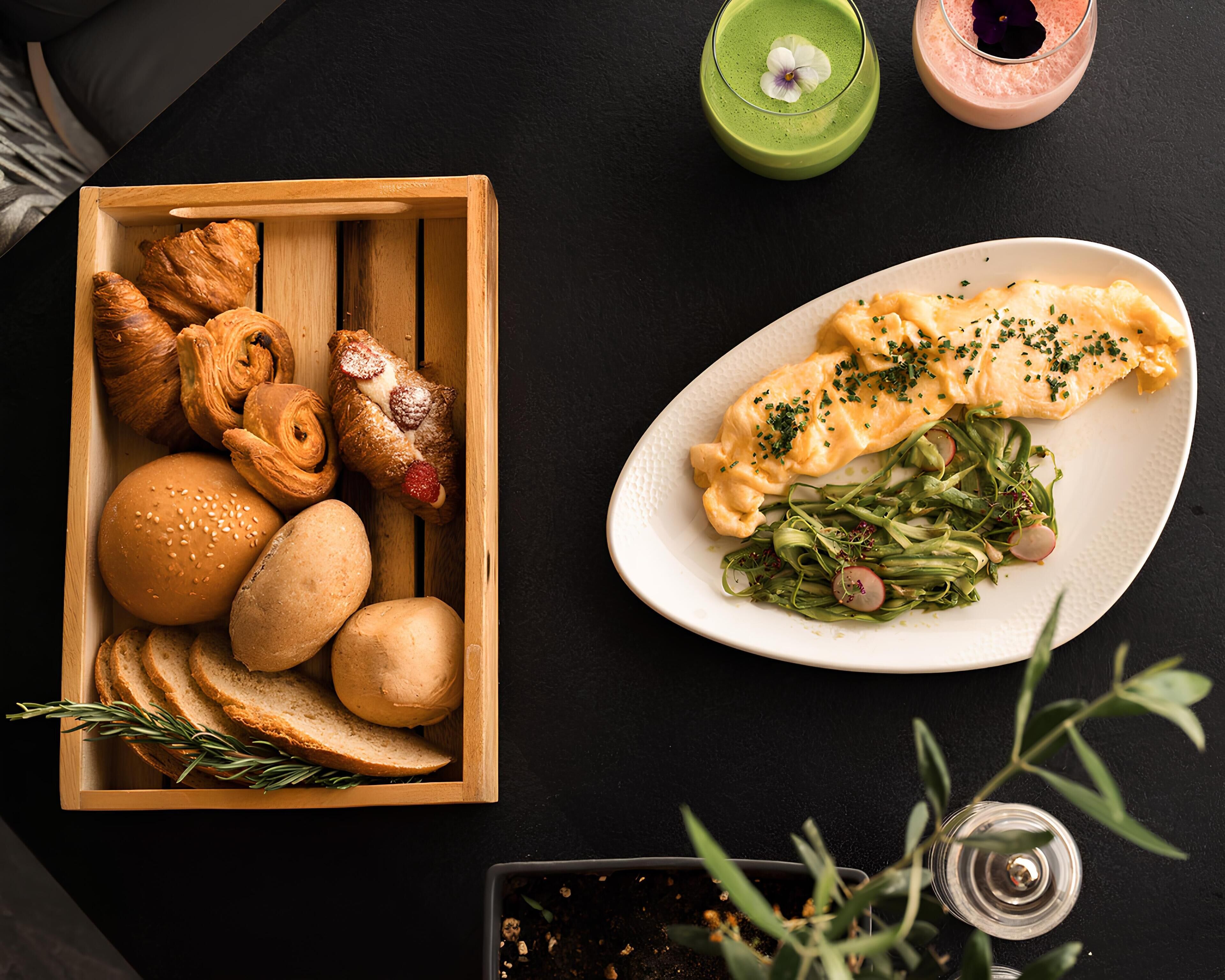 Uma bandeja de madeira com diversos pães e doces, um prato com salada verde e um ovo pochê, e duas bebidas sobre uma superfície escura.