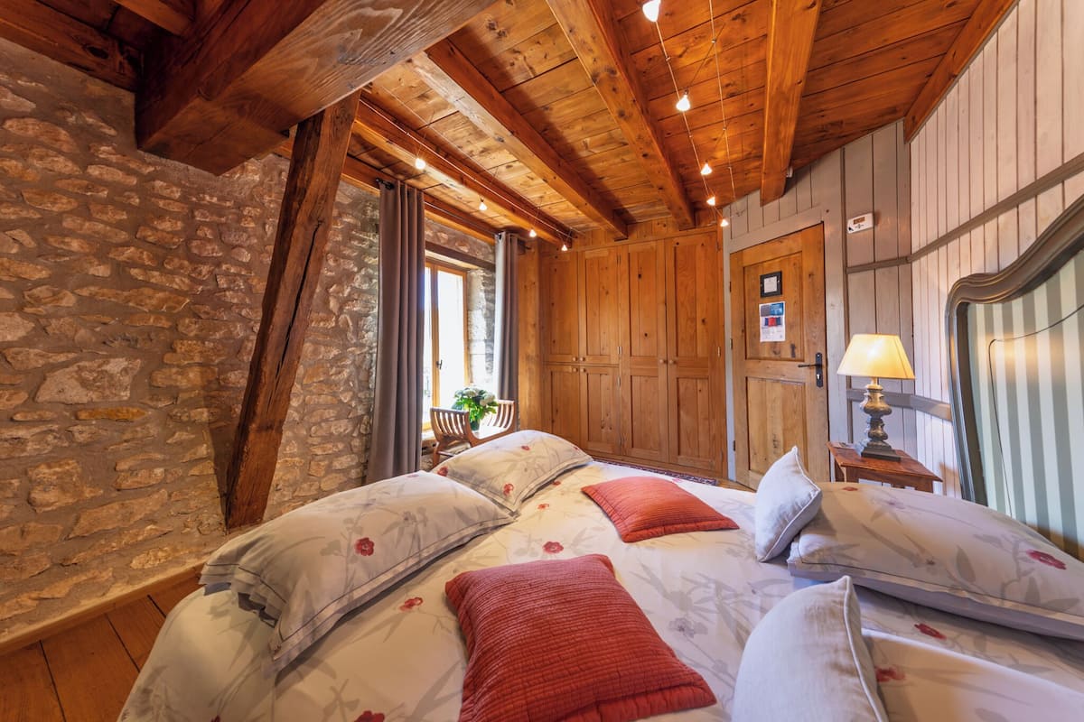 Quarto aconchegante com parede de pedra, vigas de madeira no teto e cama com roupa de cama floral e almofadas vermelhas.