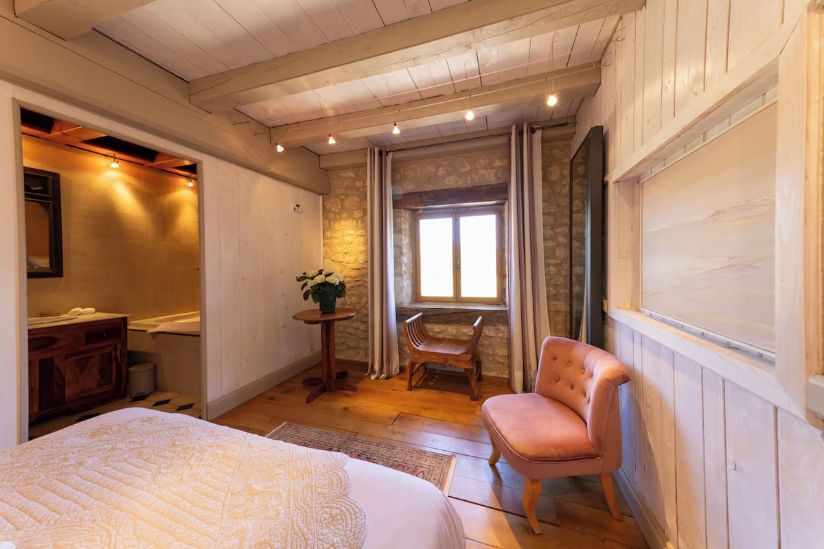 Quarto com teto de madeira, cama, poltrona, mesa pequena com planta e janela com cortinas.