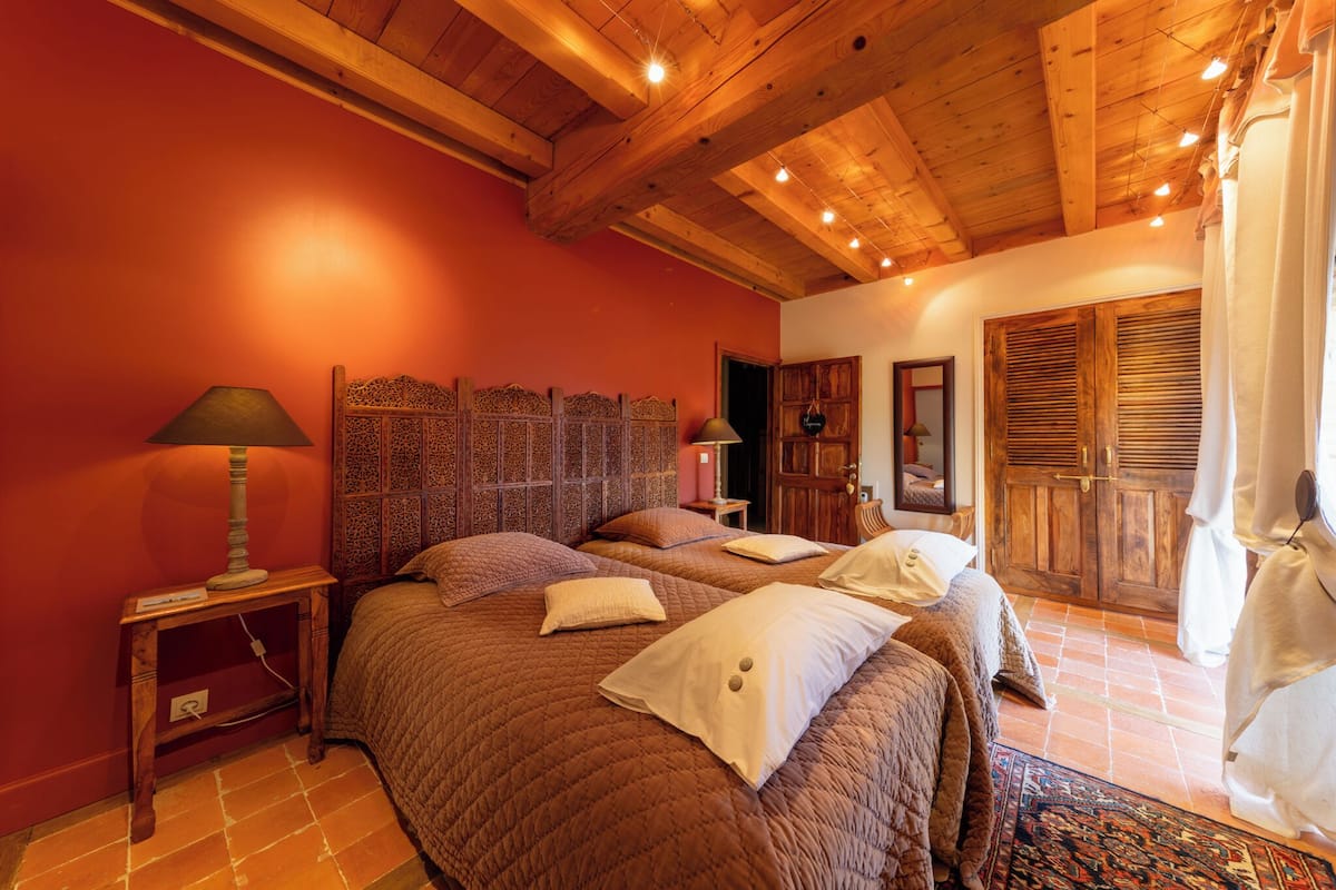 Quarto com duas camas, cabeceira de madeira, mesa de cabeceira com abajur e porta que dá para outro cômodo.