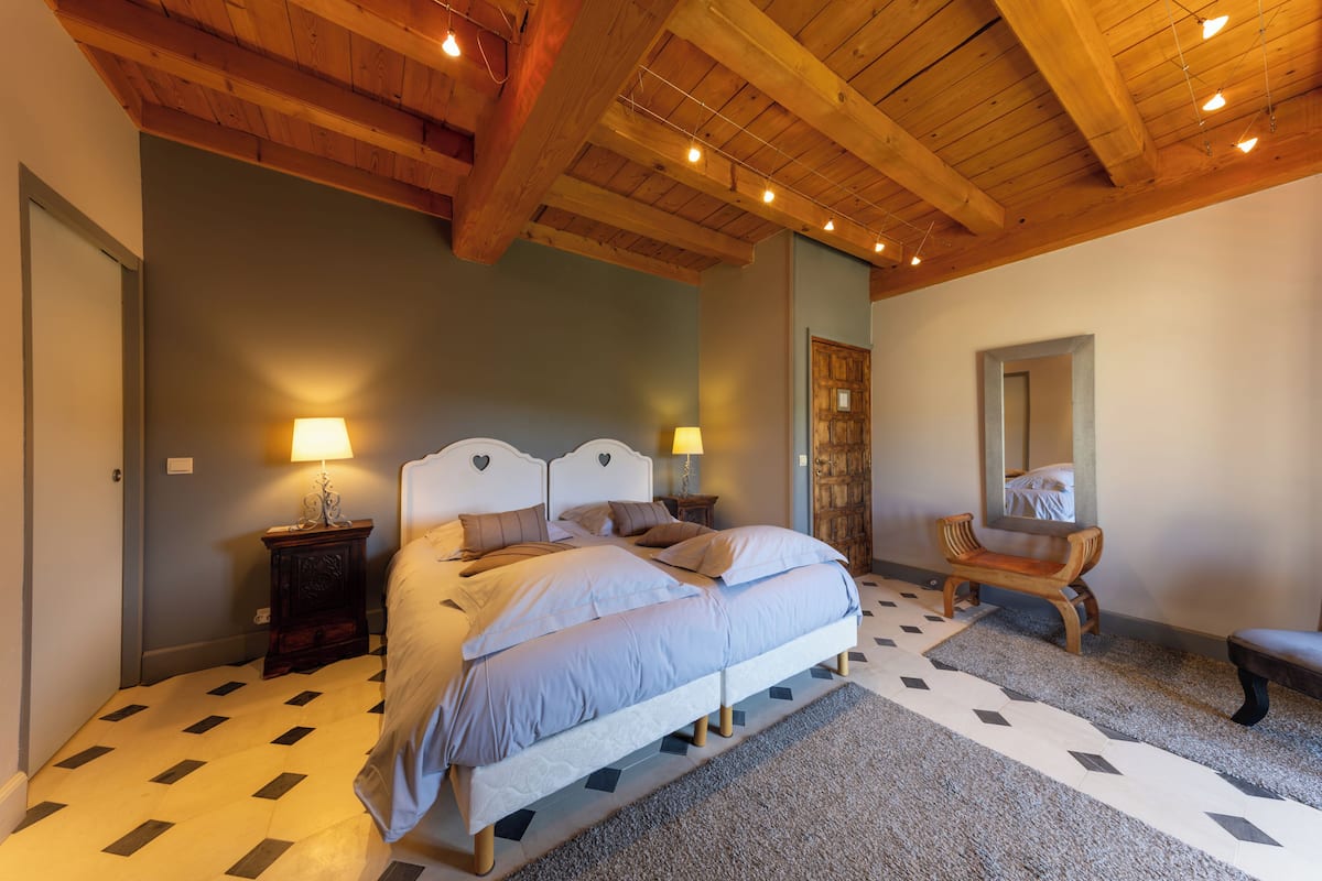 Quarto com cama, duas mesas de cabeceira, um banquinho, um espelho e um chão estampado.