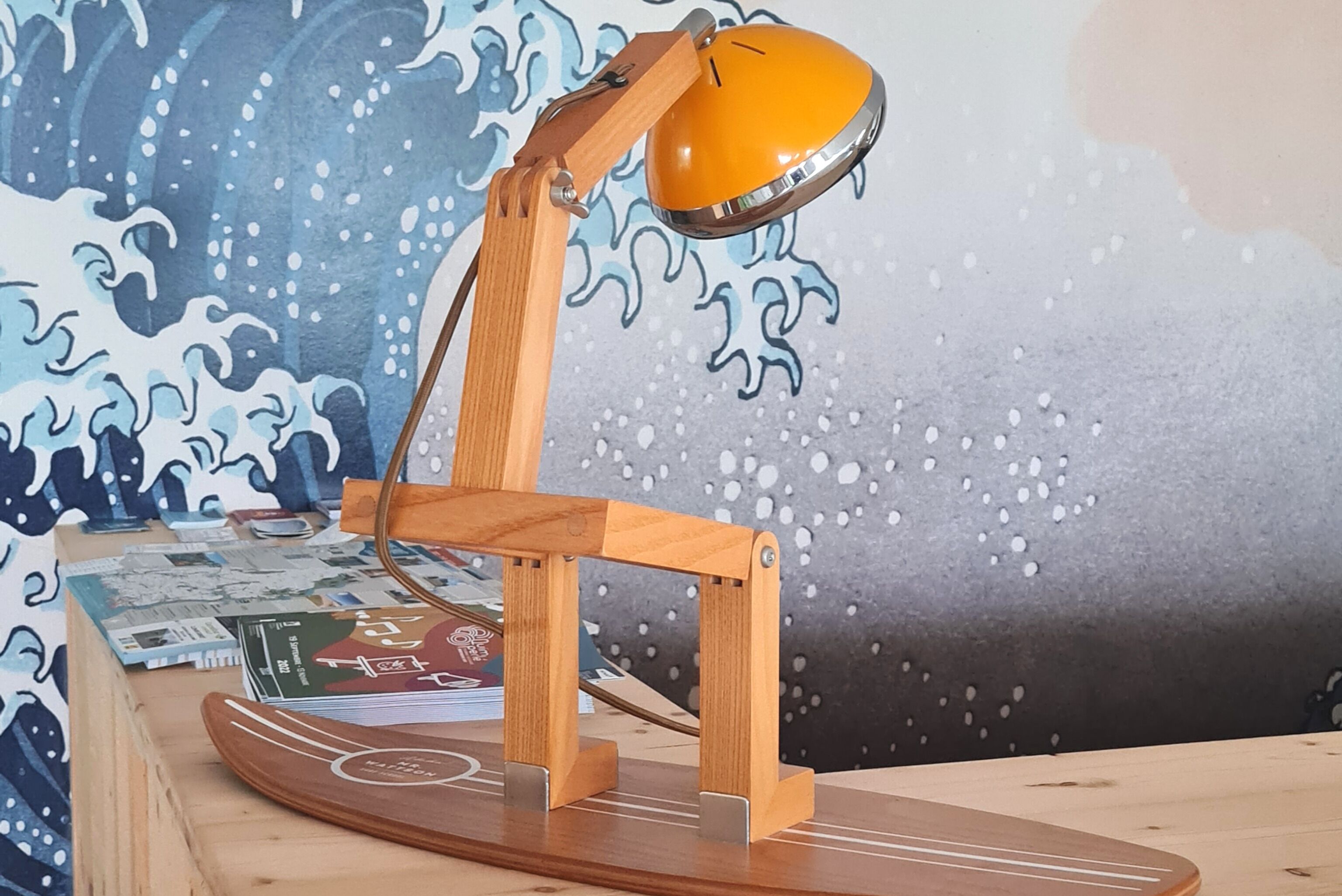 Uma escrivaninha de madeira com uma luminária amarela, base em formato de prancha de surf e parede ao fundo com padrão de ondas.