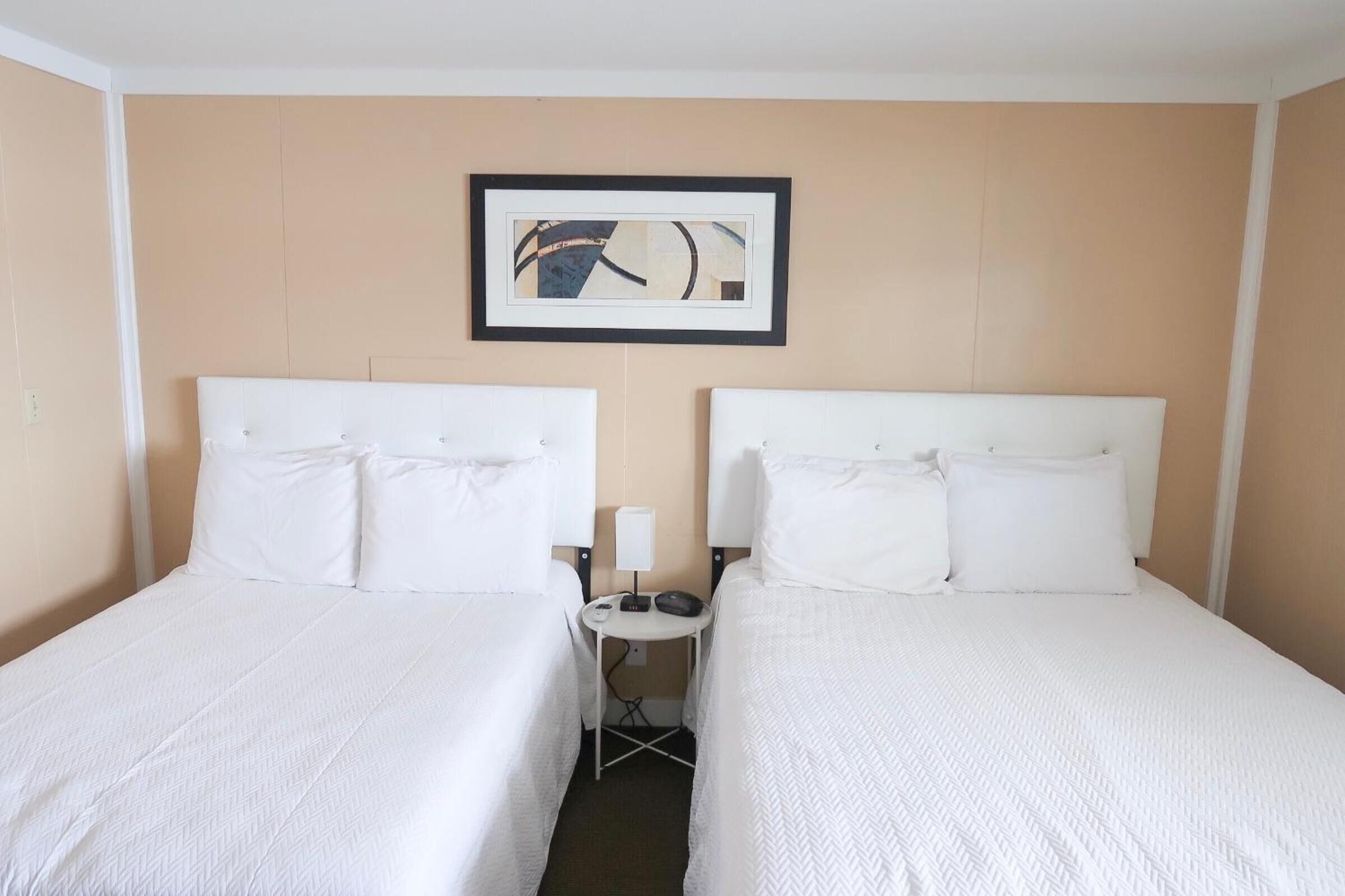 Quarto de hotel com duas camas, uma mesa pequena com abajur e um quadro abstrato emoldurado na parede.
