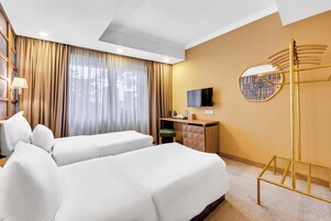 Quarto de hotel com duas camas, uma escrivaninha com televisão, uma estante de livros e uma janela grande com cortinas.