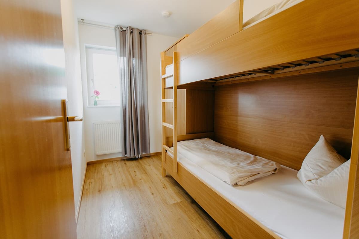 Quarto compacto com cama de solteiro, piso de madeira, radiador e janela com cortinas.