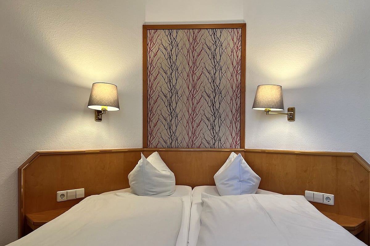 Quarto de hotel com cabeceira de madeira, duas luminárias de apoio, um separador de ambiente estampado e uma cama bem arrumada com lençóis e almofadas brancas.