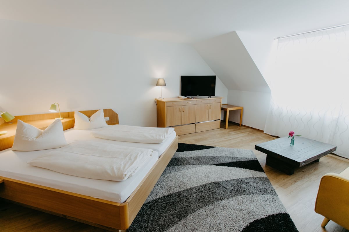 Quarto de hotel moderno com cama, escrivaninha, televisão e uma mesinha com um vaso de flores.