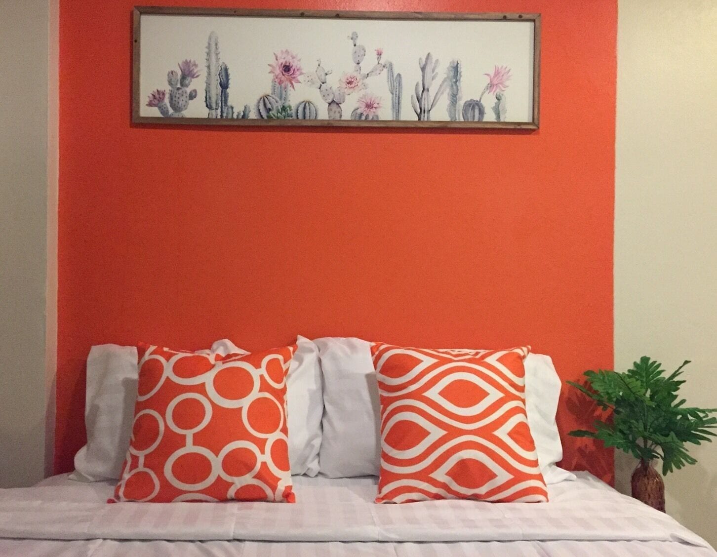 Uma cama com duas almofadas, uma obra de arte emoldurada com cactos na parede e uma planta ao lado.