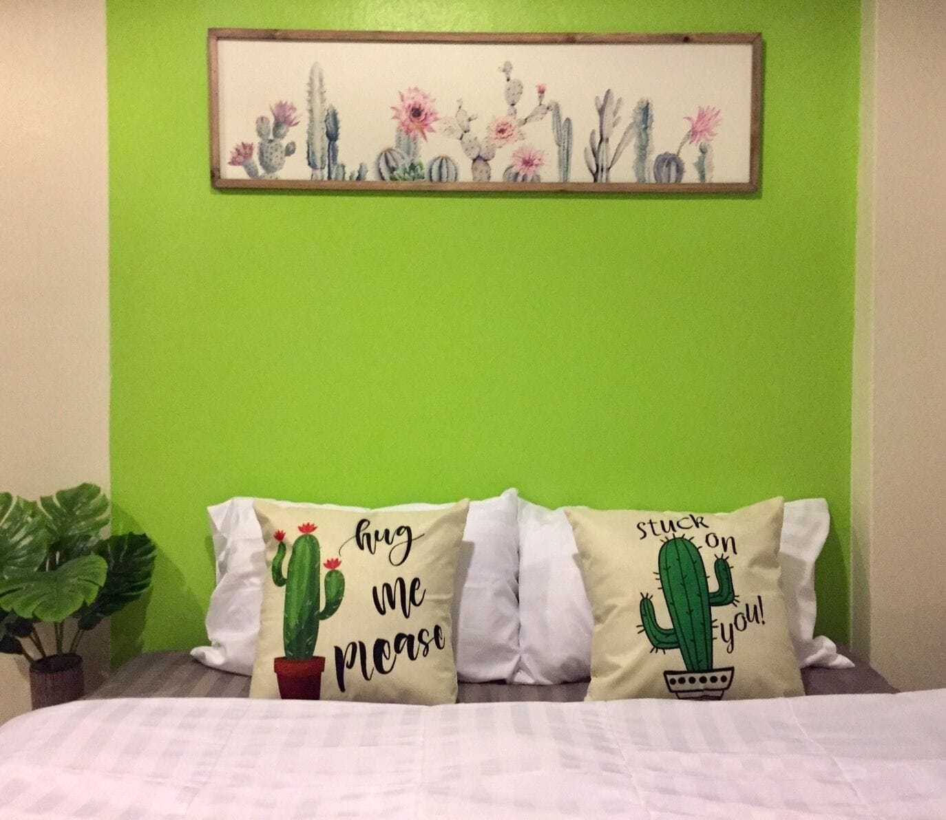 Cama com duas almofadas com estampas de cactos, uma obra de arte com cactos emoldurada em uma parede verde, e uma planta em vaso à esquerda.