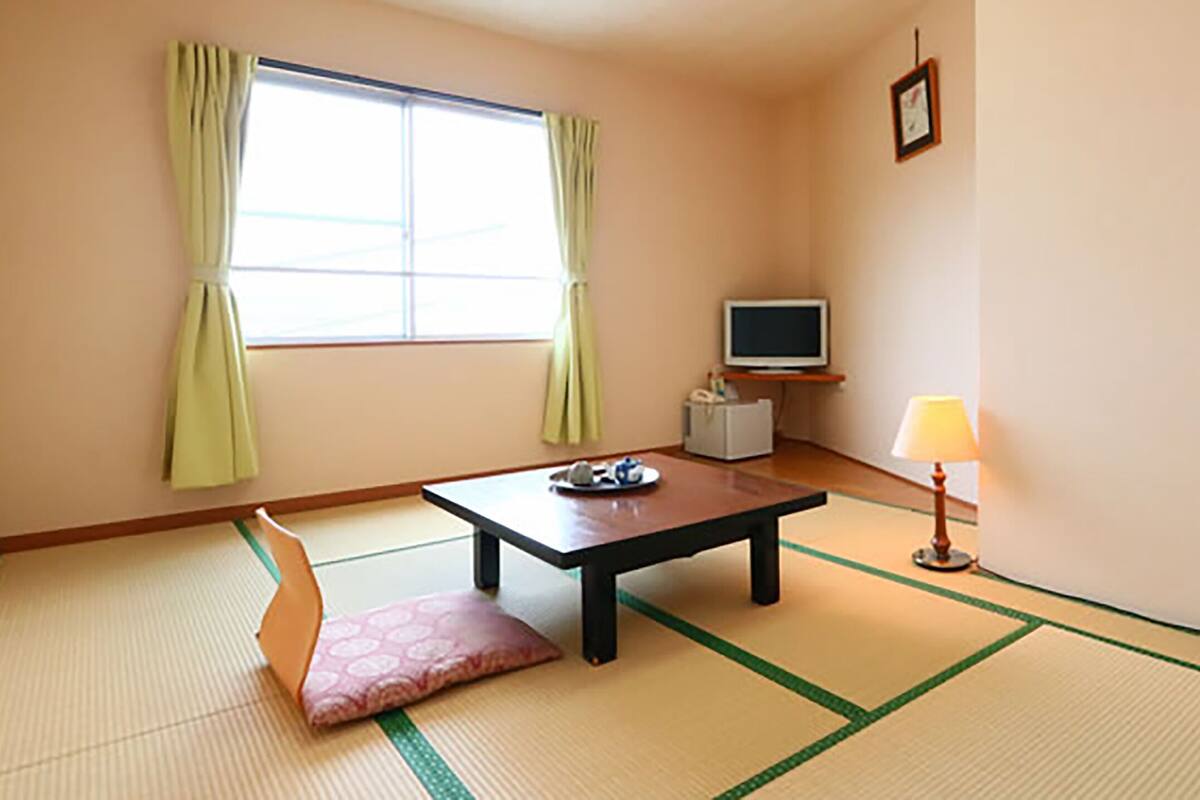Quarto com piso de tatame, uma mesa baixa, um assento almofadado, televisão e luminária.