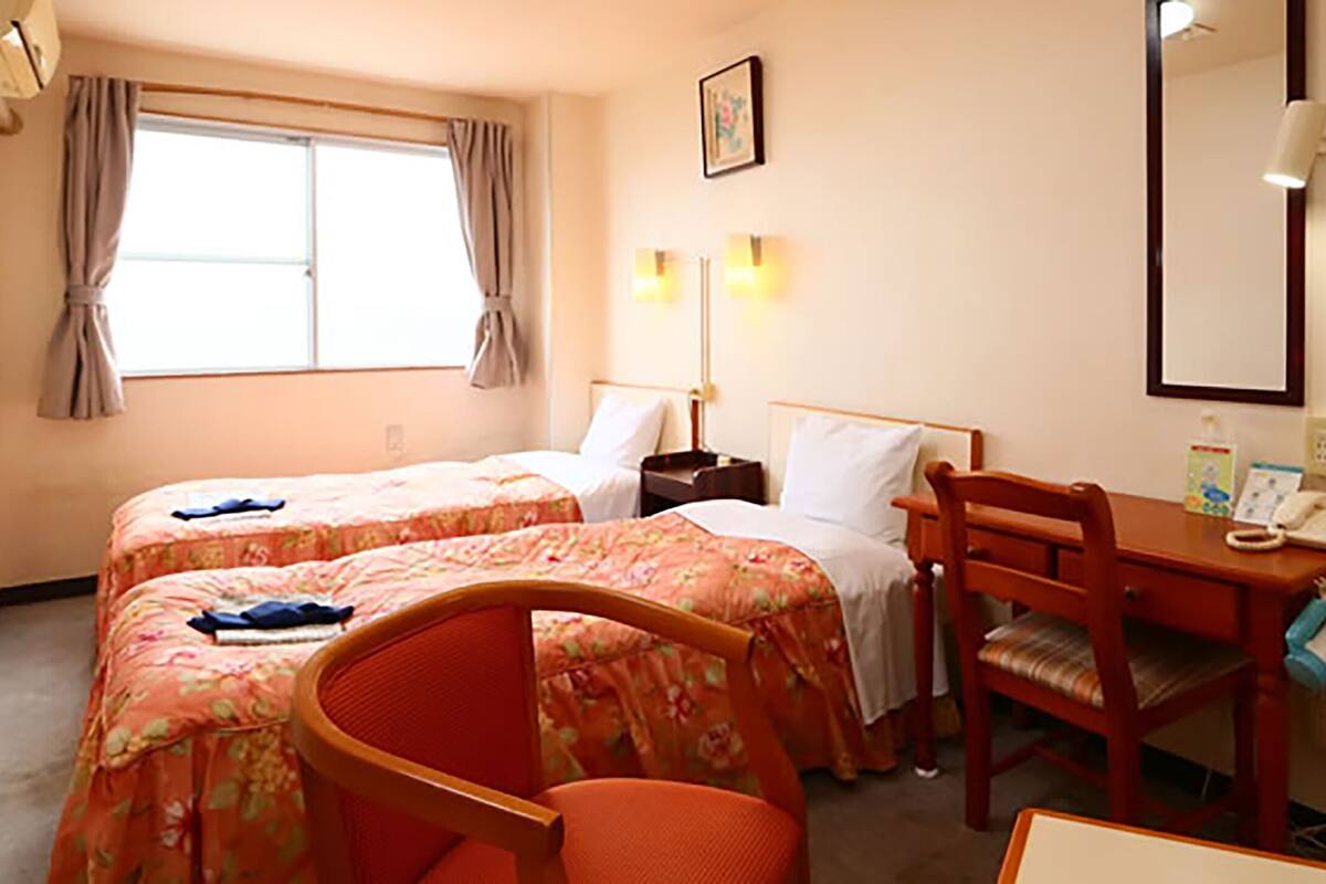 Quarto de hotel com duas camas, uma escrivaninha, uma cadeira, mesa de cabeceira, abajur, espelho e janela com cortinas.