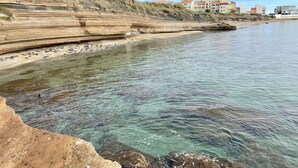Paisagem costeira com águas cristalinas, costas rochosas e edifícios ao fundo.