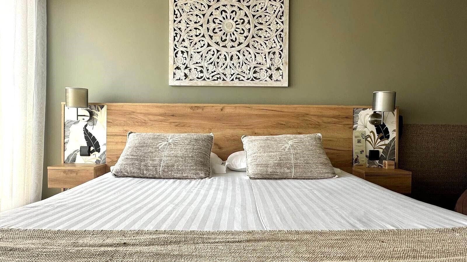 Cama bem arrumada com duas almofadas, cabeceira de madeira e um objeto decorativo na parede.