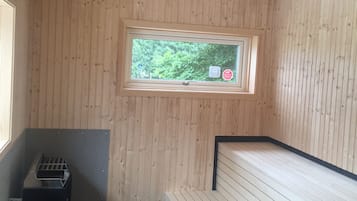 Uma cabana pequena com interior de madeira, uma janela com vista para a vegetação, um banco e um pequeno aquecedor.