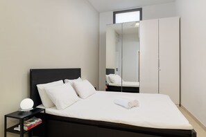Uma cama bem arrumada com roupa de cama e almofadas brancas, uma mesa de cabeceira com abajur e livros, e um guarda-roupa grande com espelho.