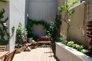 Um deck de madeira com um banco e cadeiras, cercado por plantas em vasos e uma parede de concreto.