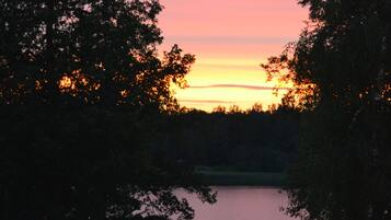 Vista serena de um lago com uma cruz em primeiro plano, rodeada por árvores e um pôr do sol ao fundo.