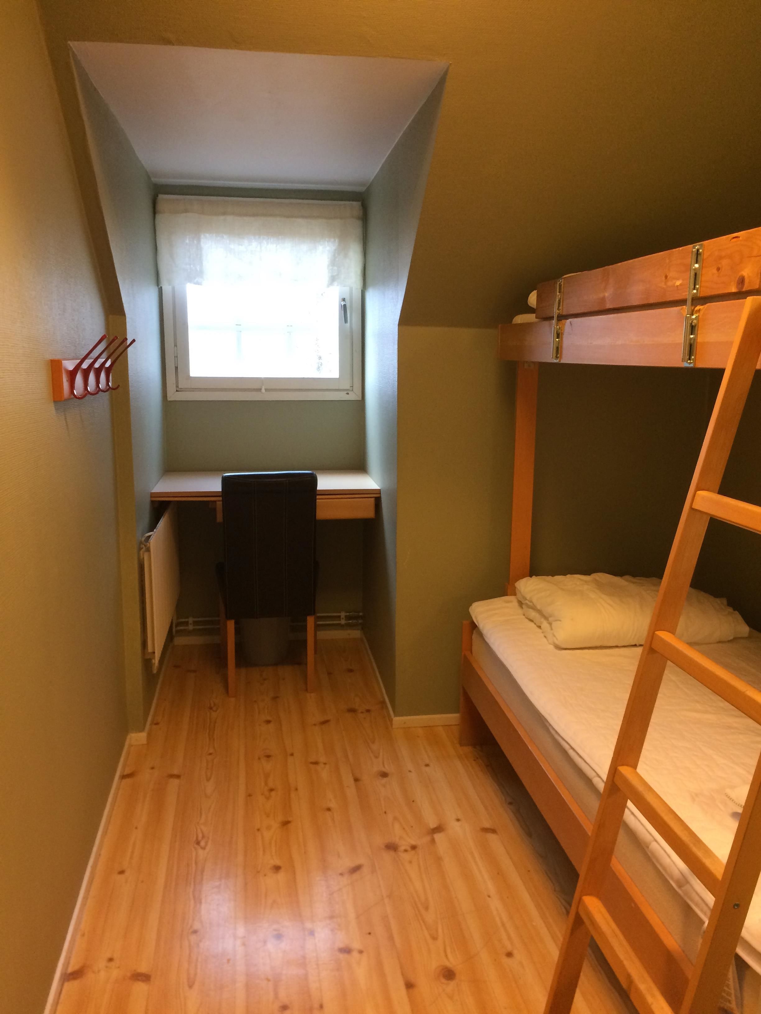 Um quarto de estudante com uma cama beliche, uma escrivaninha com cadeira e uma janela.