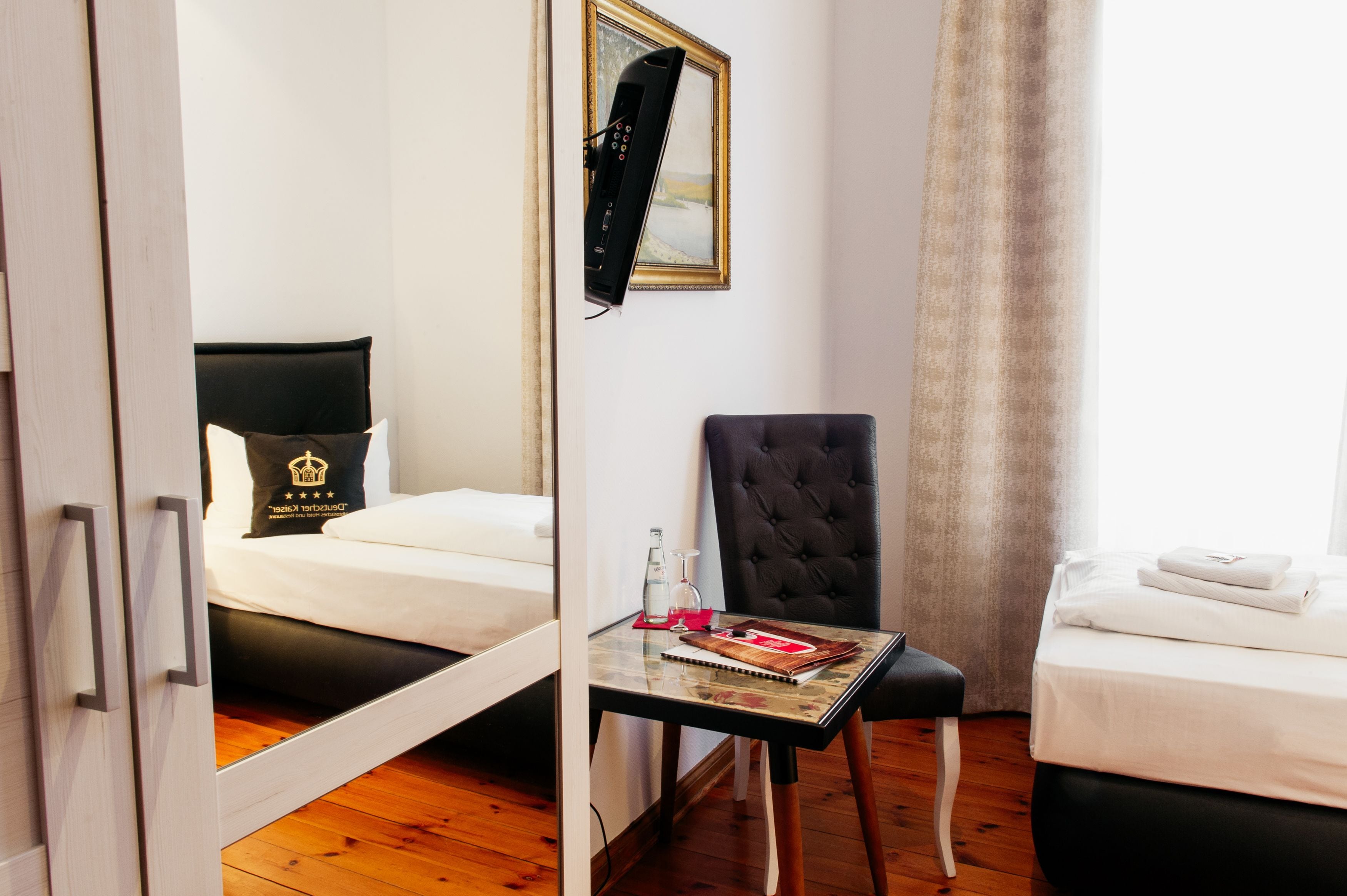 Quarto de hotel com duas camas, um espelho, uma cadeira, uma mesinha com um livro e uma garrafa, e uma janela com cortinas.