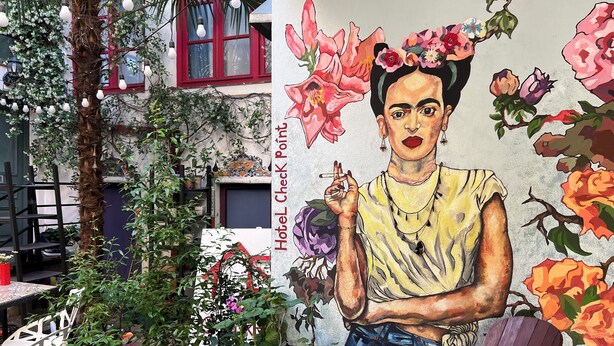 Um mural com uma pessoa usando uma coroa de flores e segurando um cigarro, uma janela vermelha e uma árvore decorada com luzes penduradas.