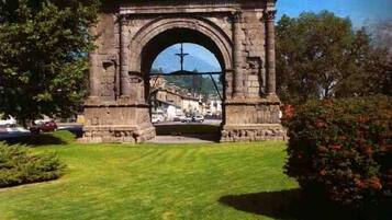 Um arco de pedra com uma estátua no centro, rodeado por vegetação e uma vista clara de uma cidade ao fundo.